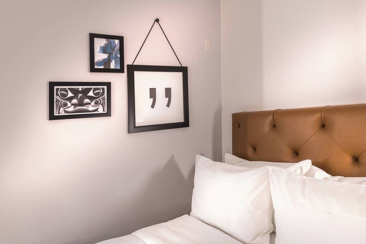 Quarto com cabeceira estofada, roupa de cama branca e três obras de arte emolduradas na parede.