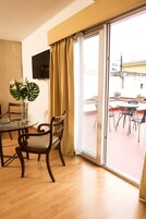 Quarto com mesa de jantar, cadeiras, TV fixada na parede e porta de vidro deslizante para um alpendre externo com cadeiras e plantas em vasos.