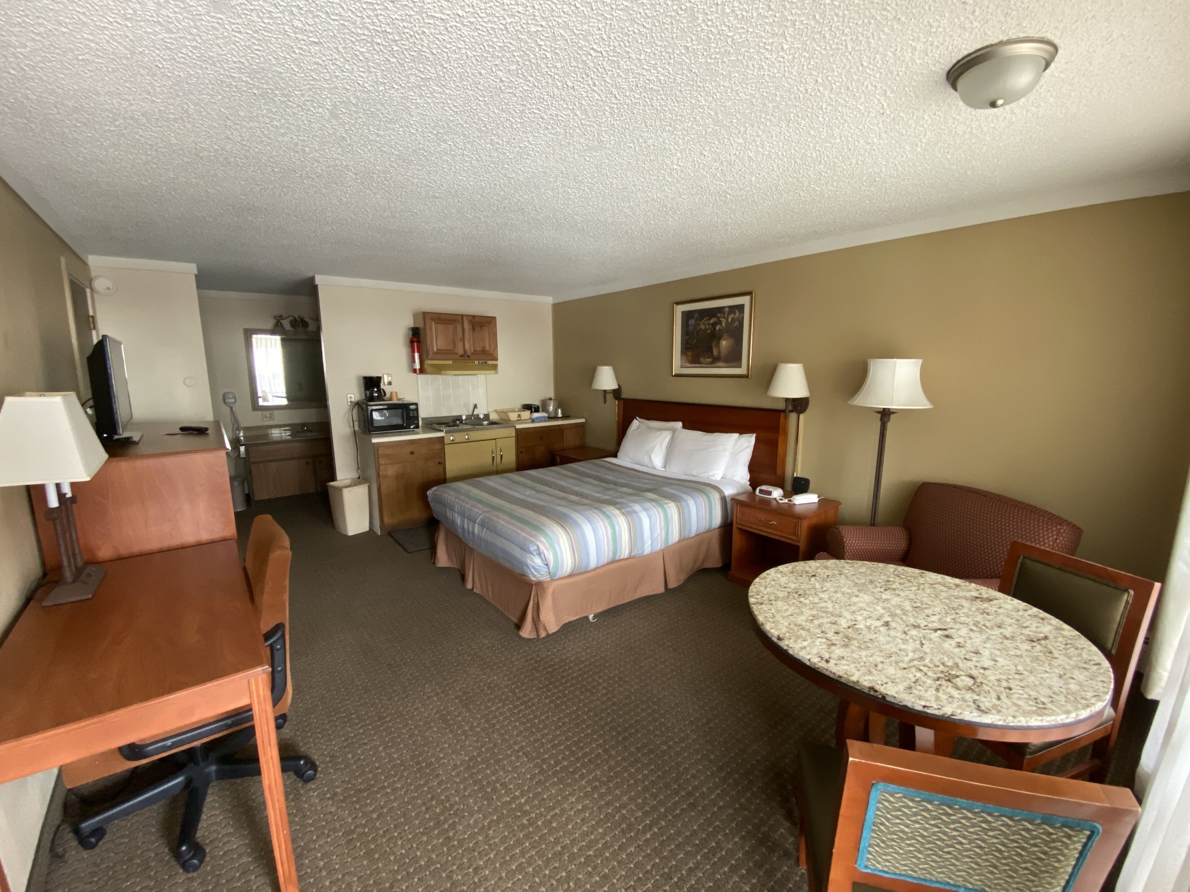 Quarto de hotel com cama, escrivaninha, cadeira, mesa de jantar e cozinha compacta.