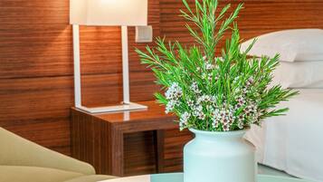 Quarto de hotel com cabeceira de madeira, mesa de cabeceira com abajur branco e um vaso com folhagem verde e flores brancas.