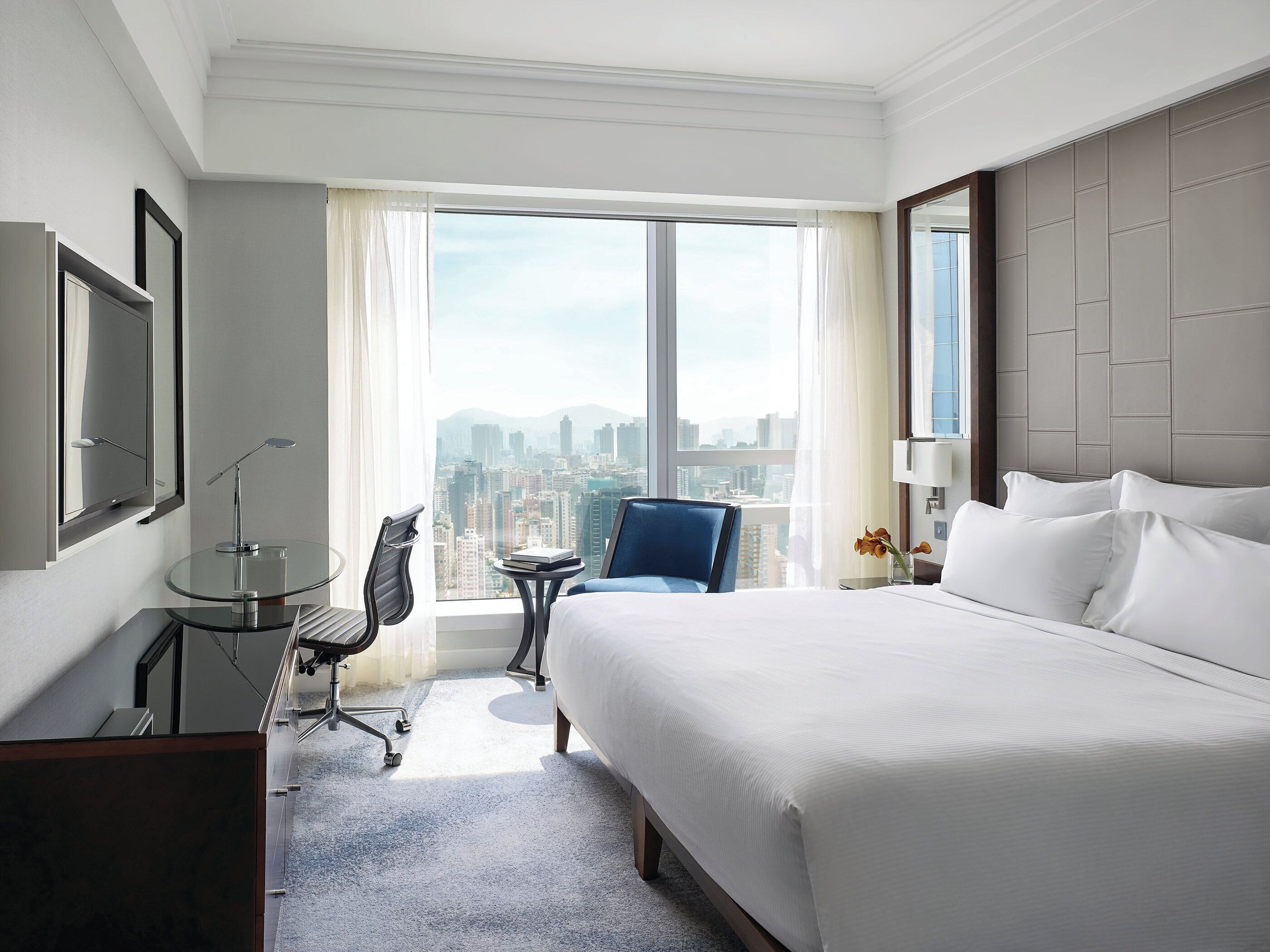 Quarto de hotel com uma cama grande, uma escrivaninha com cadeira, uma mesinha e vista para a paisagem urbana através de amplas janelas.