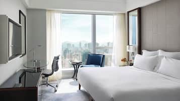 Quarto de hotel com uma cama grande, uma escrivaninha com cadeira, uma mesinha e vista para a paisagem urbana através de amplas janelas.