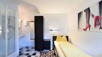 Quarto de hotel moderno com piso xadrez, cama com almofadas amarelas, mesa de cabeceira, escrivaninha e box de vidro.