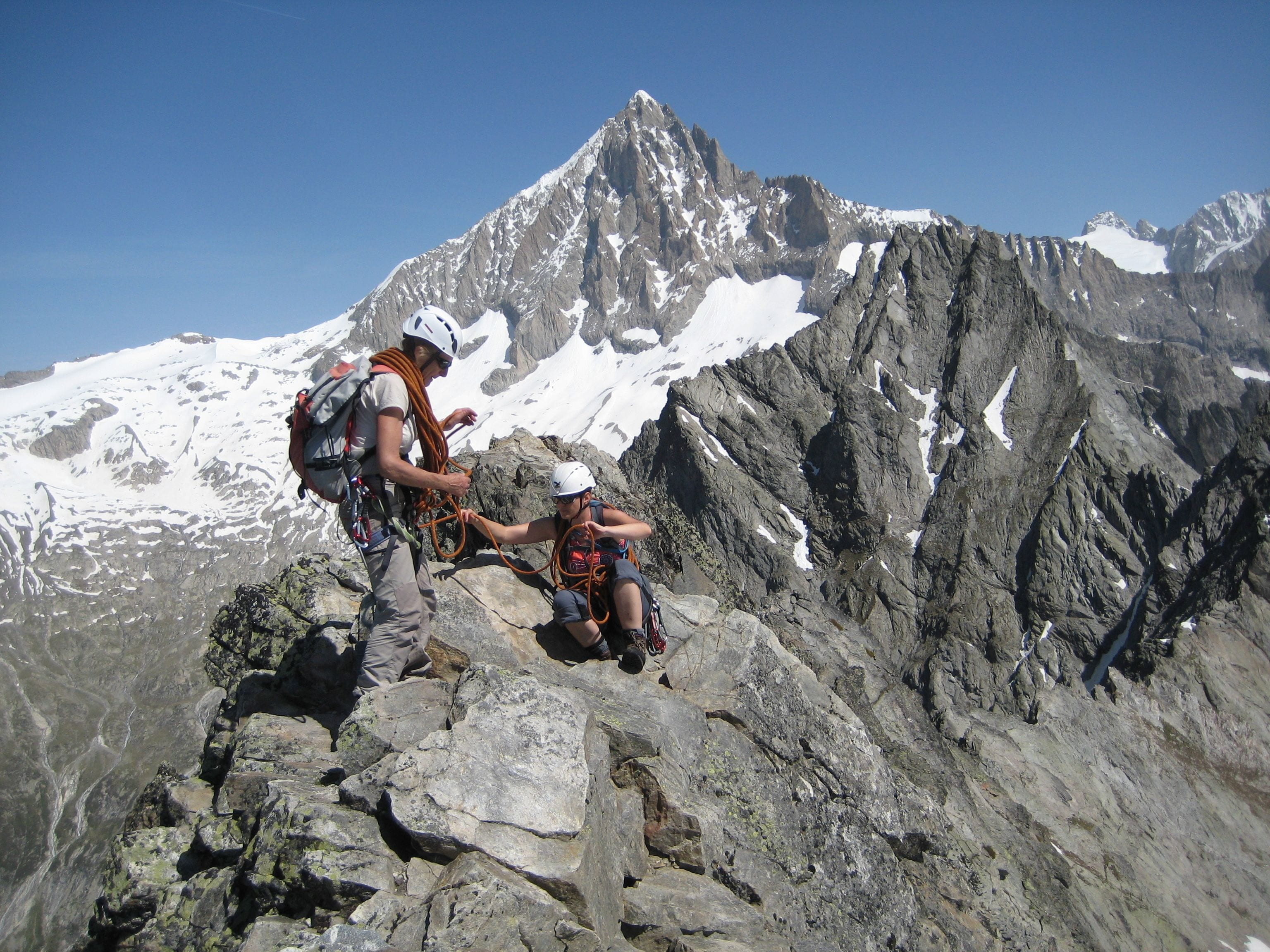 Dois alpinistas no topo de uma montanha rochosa, com neve e céu limpo.