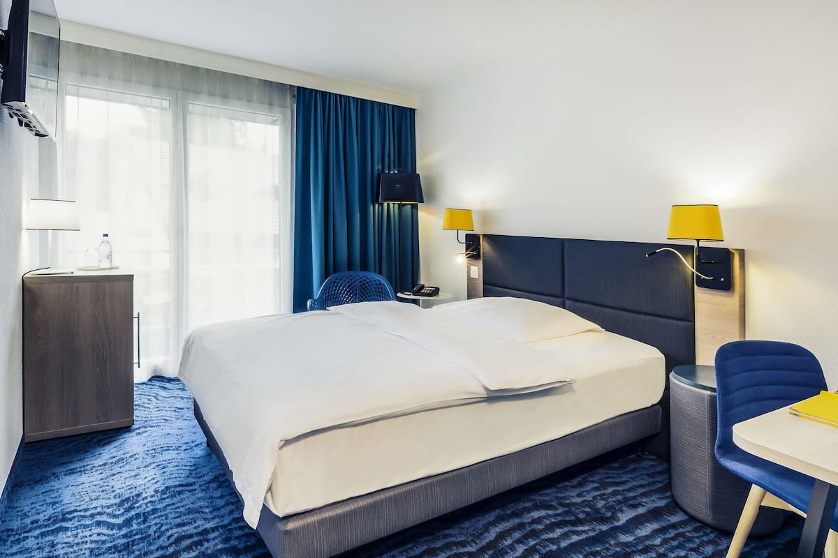 Quarto de hotel com cama, escrivaninha, cadeira, luminária e telefone em uma mesinha de cabeceira.