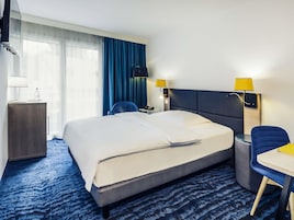 Quarto de hotel com cama, escrivaninha, cadeira, luminária e telefone em uma mesinha de cabeceira.