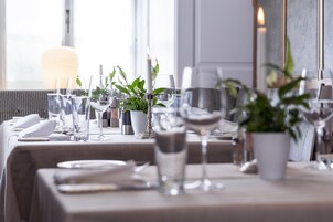 Mesa de jantar bem posta, com copos transparentes, uma vela acesa e plantas em vasos.
