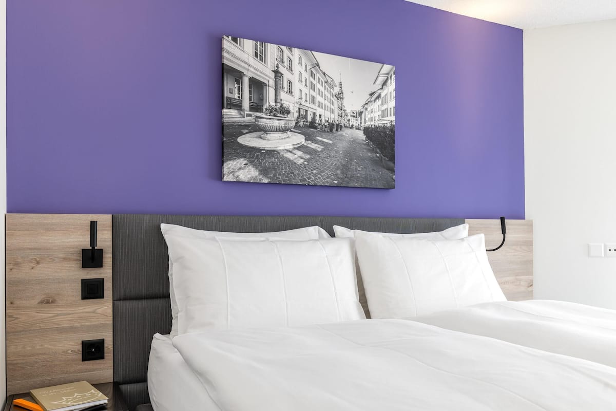 Uma cama bem arrumada com lençóis brancos, cabeceira com painel cinza e uma fotografia preto e branco de uma cena urbana na parede.
