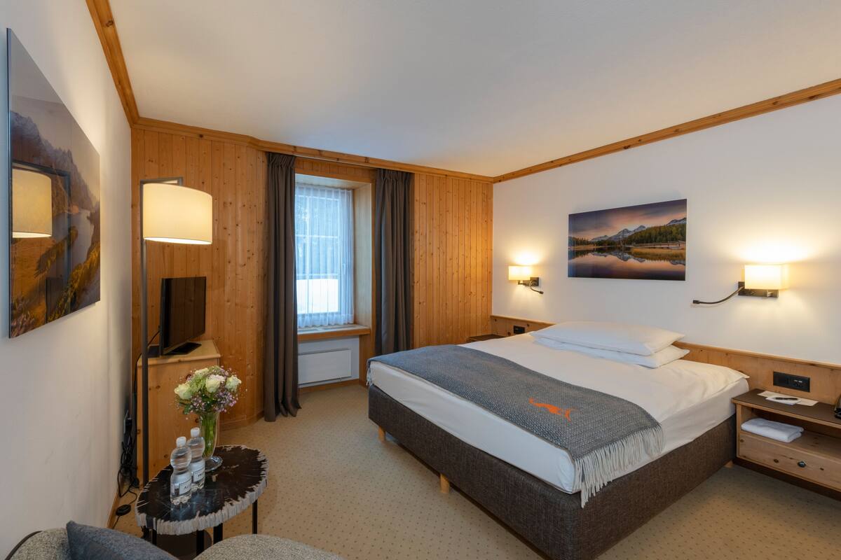 Quarto de hotel com uma cama grande, uma escrivaninha com luminária, uma TV e um quadro na parede.