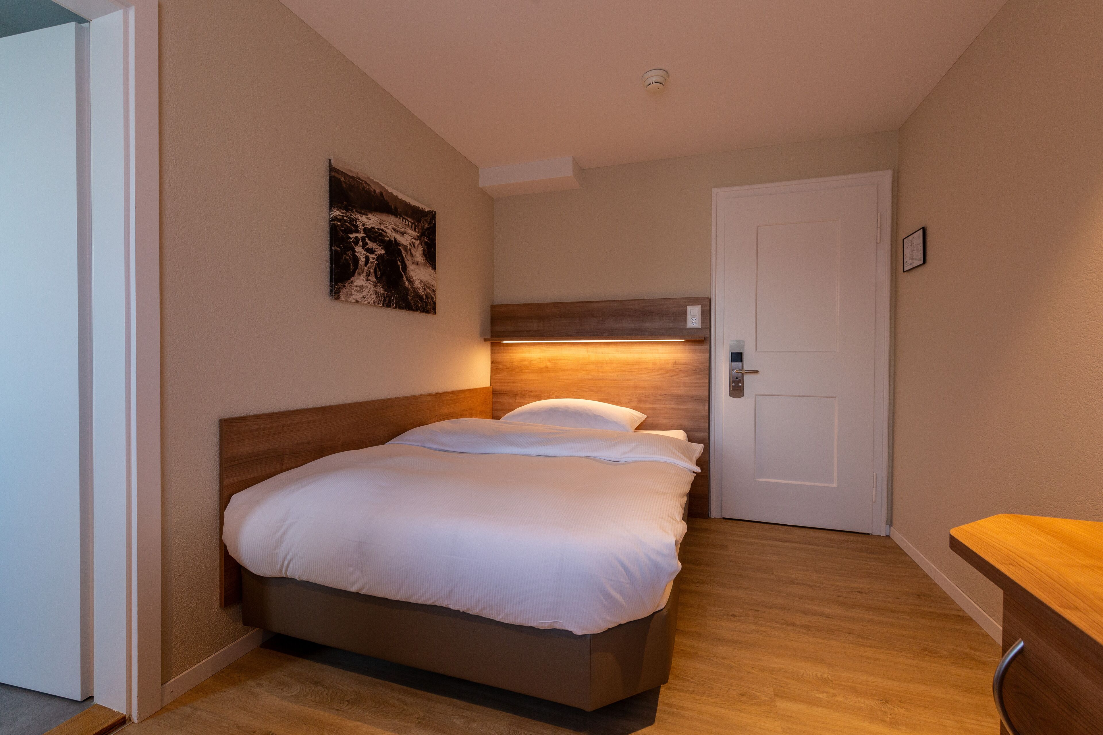 Quarto de hotel moderno com cabeceira de madeira, cama branca, pequena mesa de cabeceira e porta com puxador.