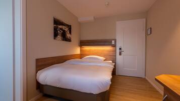 Quarto de hotel moderno com cabeceira de madeira, cama branca, pequena mesa de cabeceira e porta com puxador.