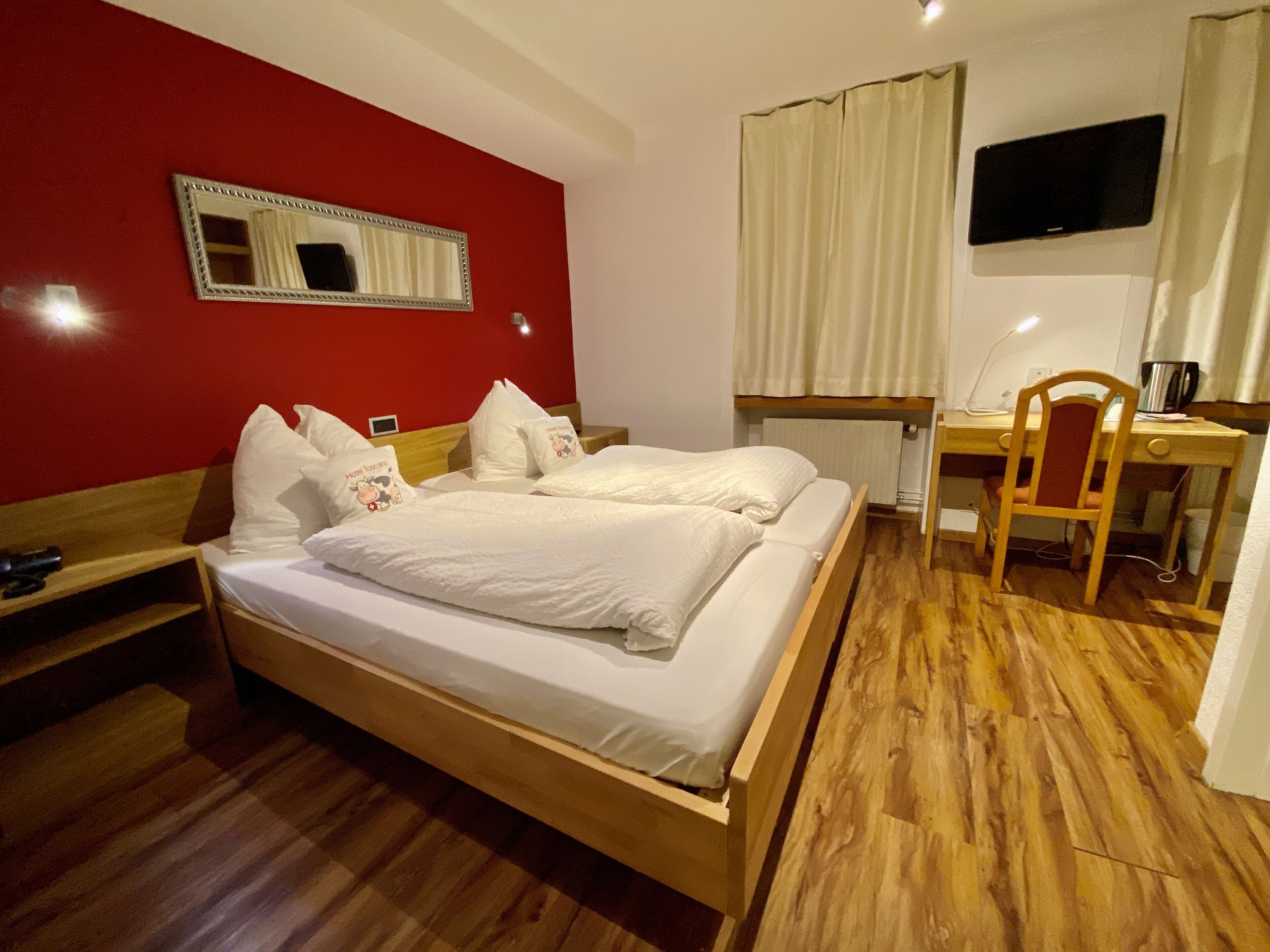 Quarto de hotel com cama de casal, escrivaninha de madeira, uma cadeira, televisão e parede vermelha com espelho.