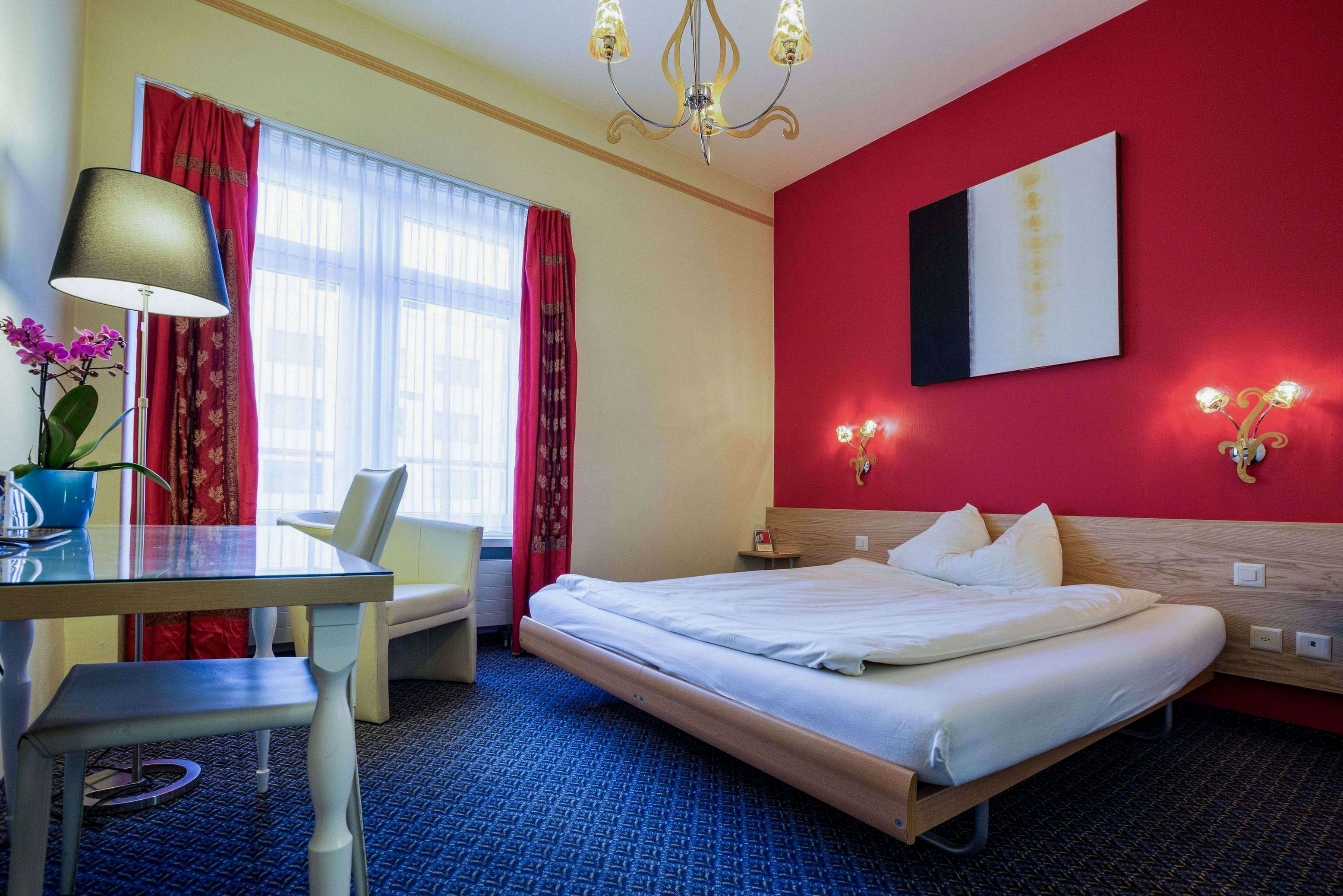 Quarto de hotel com cama, escrivaninha, cadeira, abajur, vaso com flores e parede vermelha com um quadro.