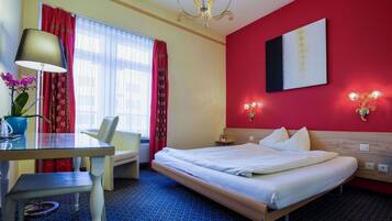 Quarto de hotel com cama, escrivaninha, cadeira, abajur, vaso com flores e parede vermelha com um quadro.