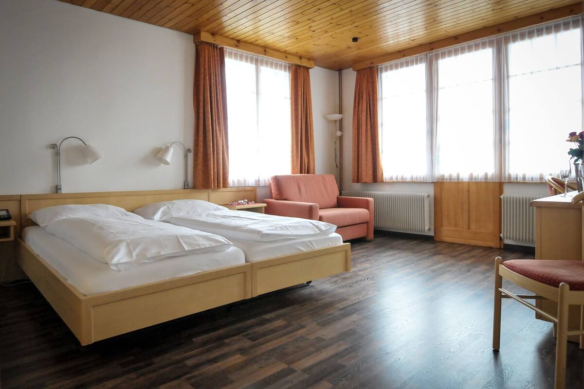 Quarto com teto de madeira, duas camas, uma cadeira, escrivaninha e janelas amplas com cortinas.