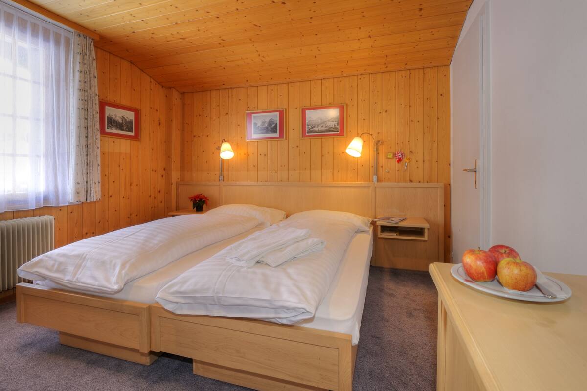 Quarto com painéis de madeira, duas camas, quadros nas paredes e uma mesa de cabeceira com maçãs.