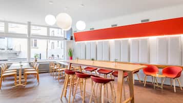 Um restaurante moderno com uma longa mesa de madeira e cadeiras vermelhas, janelas amplas e uma parede de destaque vermelha.