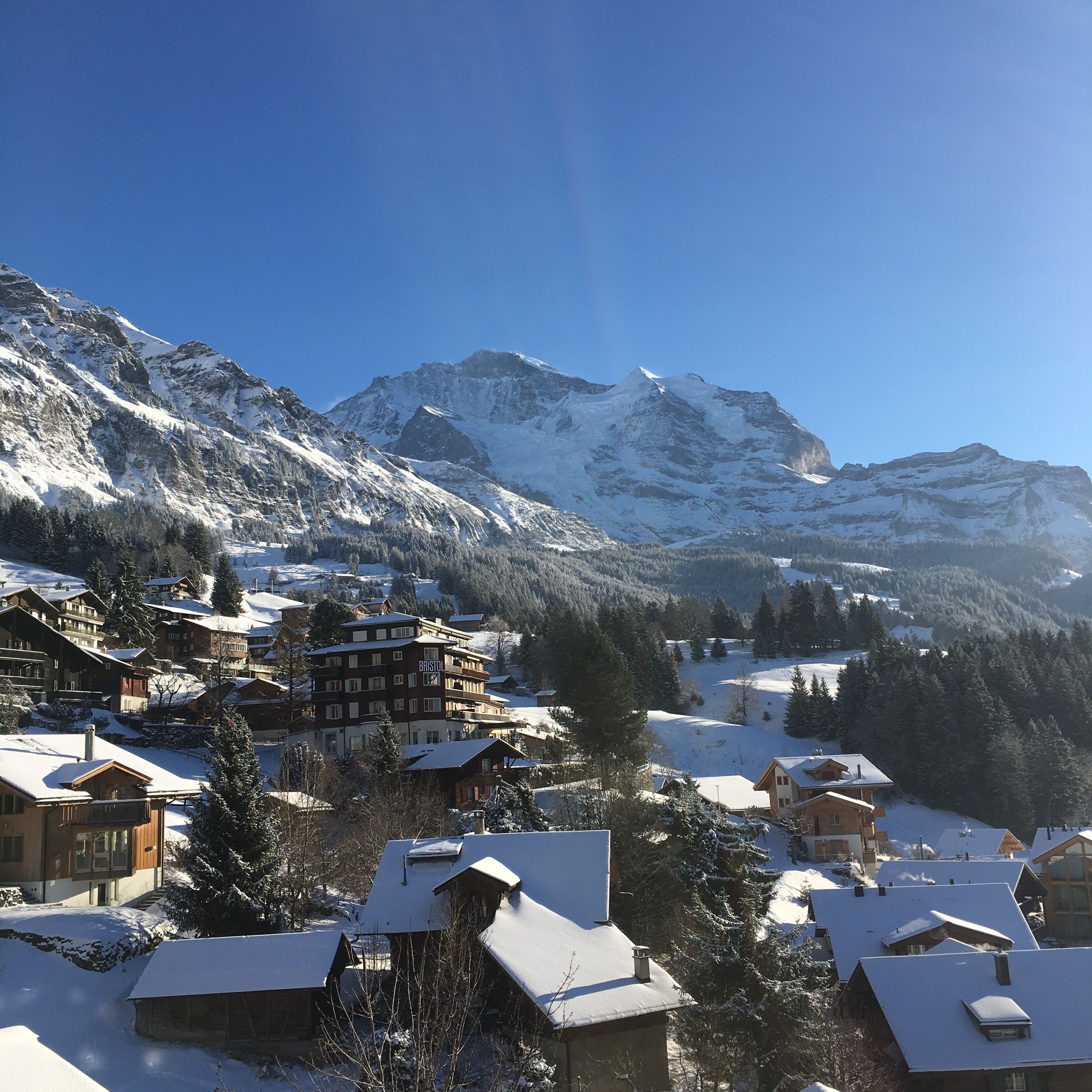 Uma aldeia montanhosa coberta de neve, com edifícios em estilo chalé, um hotel em destaque chamado 'BRISTOL', e um céu azul límpido.