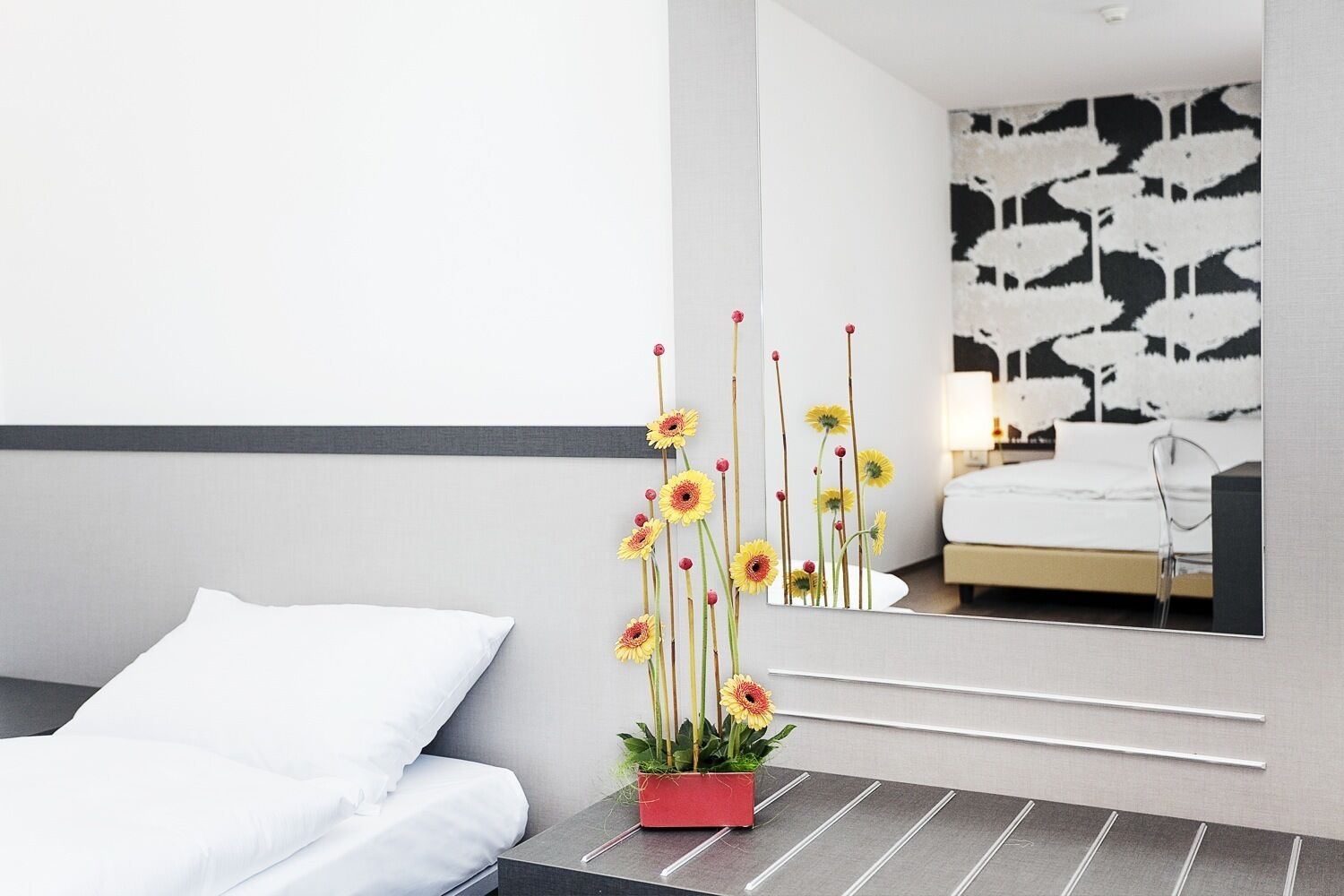Quarto de hotel moderno com uma cama, uma mesa de cabeceira com um vaso de flores vermelho, um espelho refletindo o quarto e uma parede com padrão preto e branco.