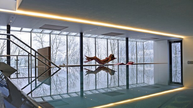 Piscina interior moderna com parede de vidro, escada de metal e design minimalista.