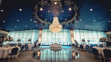 Um espaço para eventos formais com mesas redondas, cadeiras, palco central e um grande candelabro.