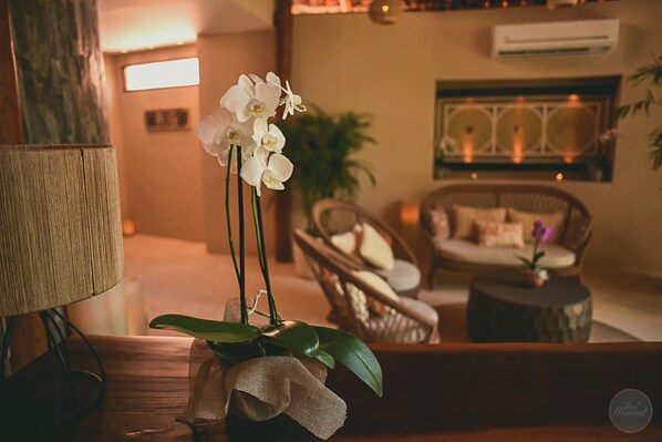 Um quarto com uma luminária, uma cadeira de vime, uma mesinha e uma orquídea em vaso.