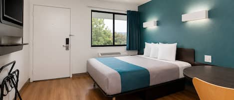 Quarto de hotel com uma cama, escrivaninha, cadeira, televisão e janela com cortinas.