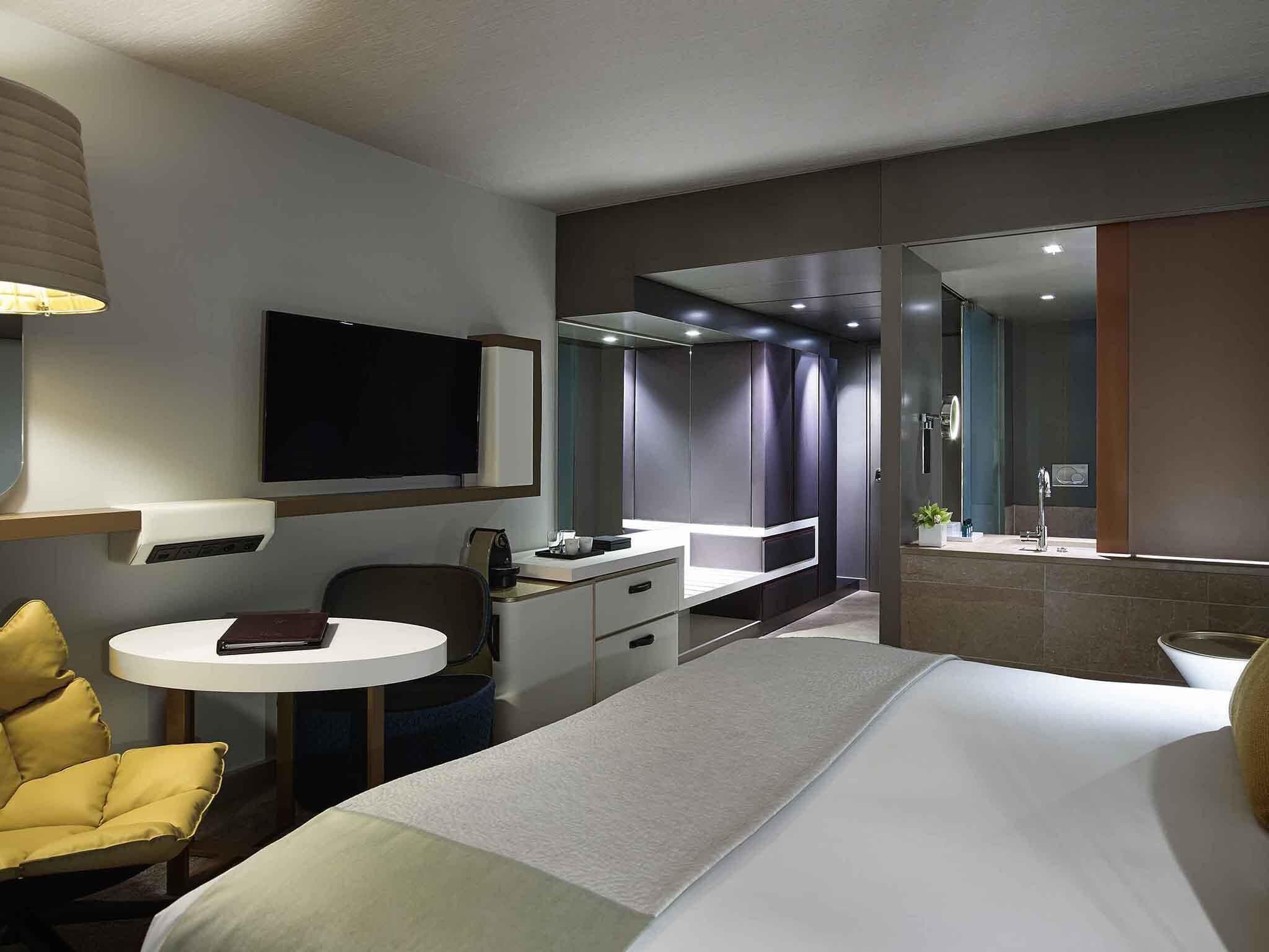 Quarto de hotel com cama, escrivaninha, cadeira, televisão, espelho e banheiro.
