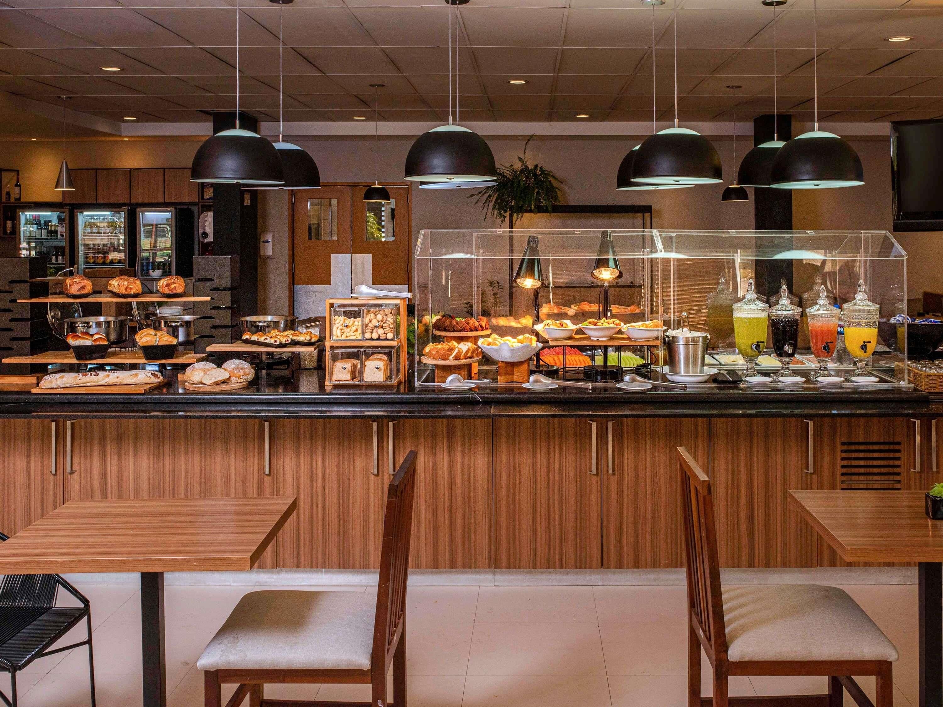Um buffet de café da manhã com diversas opções de alimentos e bebidas, móveis de madeira e luminárias pendentes.