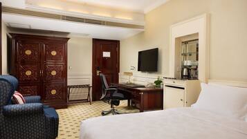 Quarto de hotel com cama, escrivaninha, cadeira, televisão, guarda-roupa e espelho.