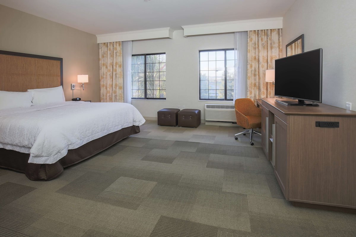 Quarto de hotel com uma cama grande, uma escrivaninha com televisão, dois pufes marrons e uma cadeira.