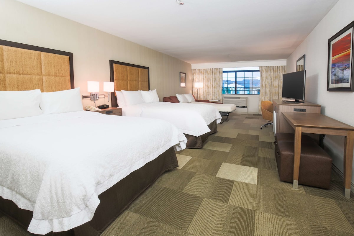 Quarto de hotel com duas camas, uma escrivaninha, uma cadeira, televisão e janela com vista.