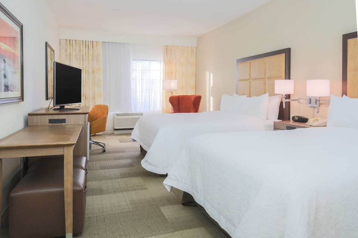 Quarto de hotel com duas camas, uma escrivaninha com computador, uma cadeira e uma televisão.