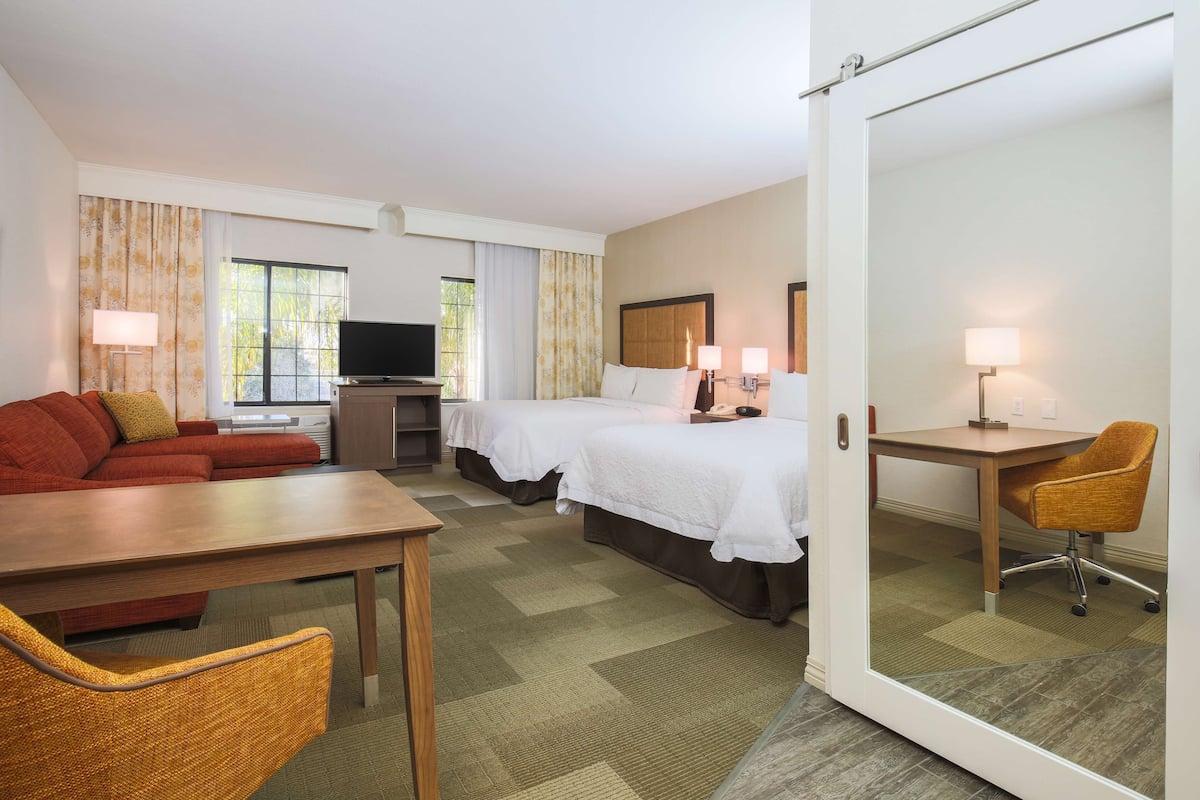 Quarto de hotel com sofá, duas camas, escrivaninha, televisão e uma cadeira.