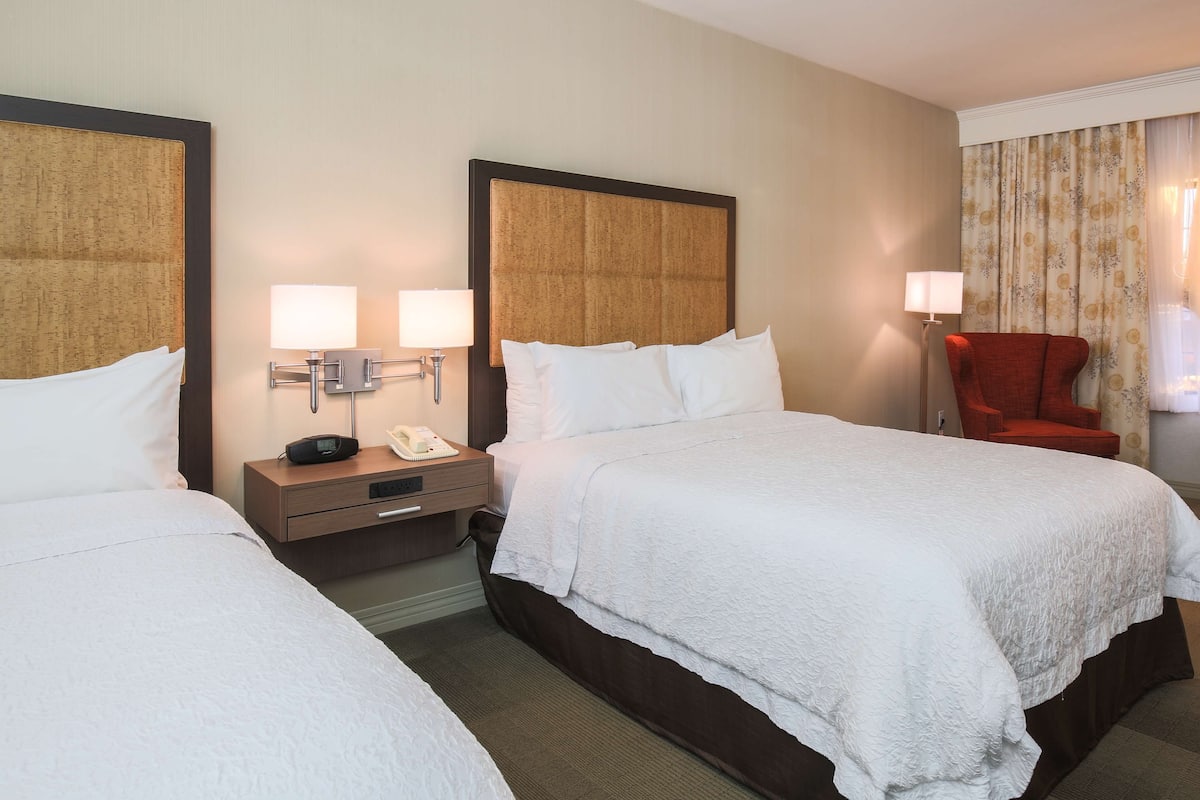 Quarto de hotel com duas camas, uma mesinha de cabeceira com abajur, uma cadeira e uma janela com cortinas.