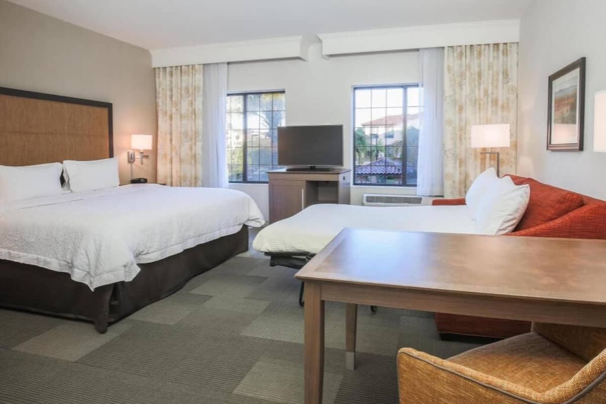 Quarto de hotel com cama, escrivaninha, televisão e sofá.