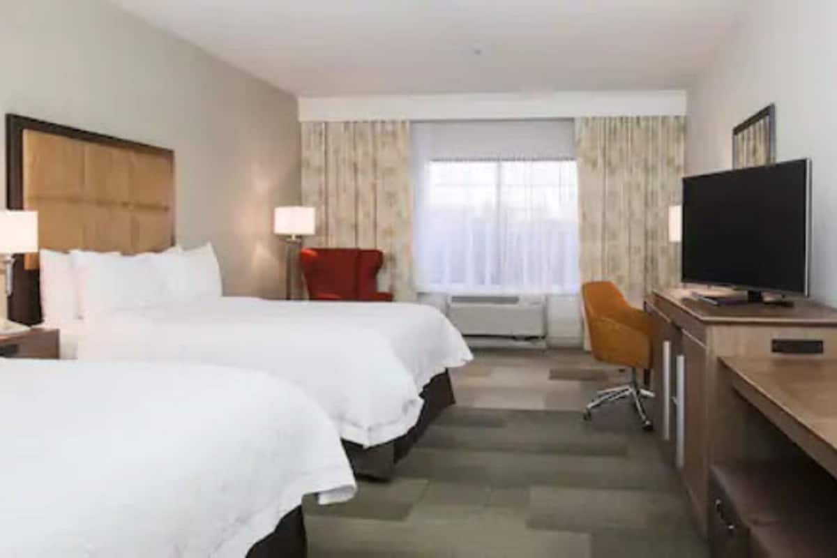 Quarto de hotel com duas camas, uma mesa de trabalho com televisão, uma cadeira e uma janela com cortinas.