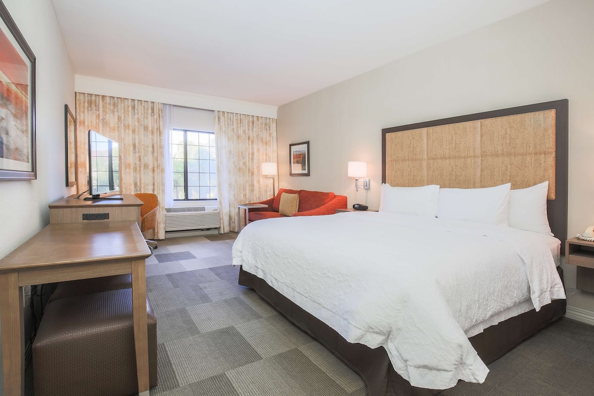 Quarto de hotel com uma cama grande, uma escrivaninha, uma cadeira e uma janela com cortinas.