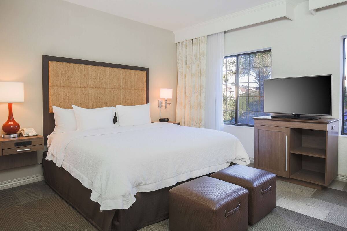 Quarto de hotel com cama grande, televisão, escrivaninha e janela com cortinas.