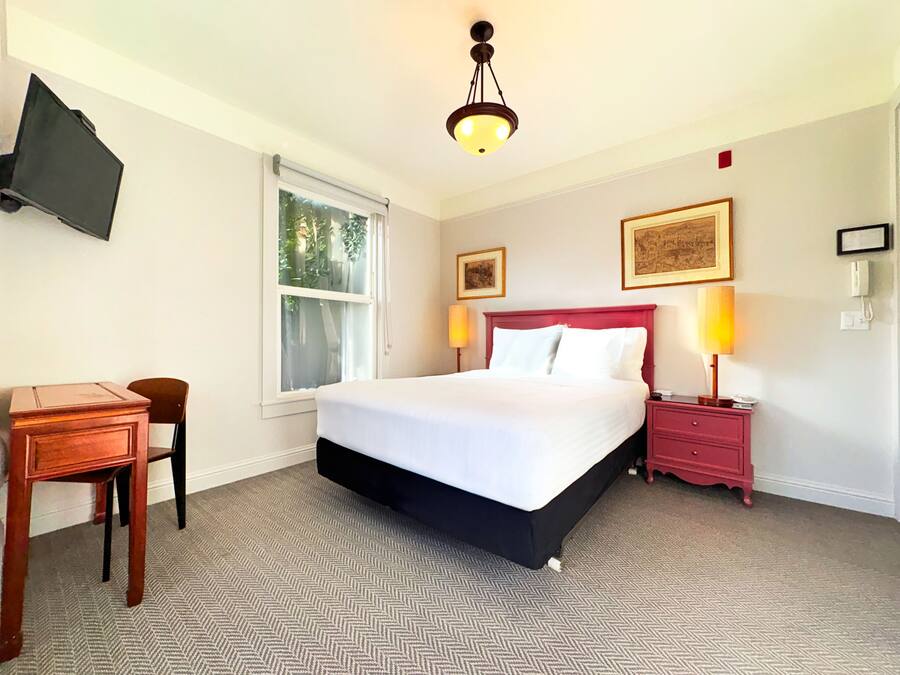 Quarto de hotel com cama, mesas de cabeceira, escrivaninha, cadeira, TV fixada na parede e janela com vista para vegetação.