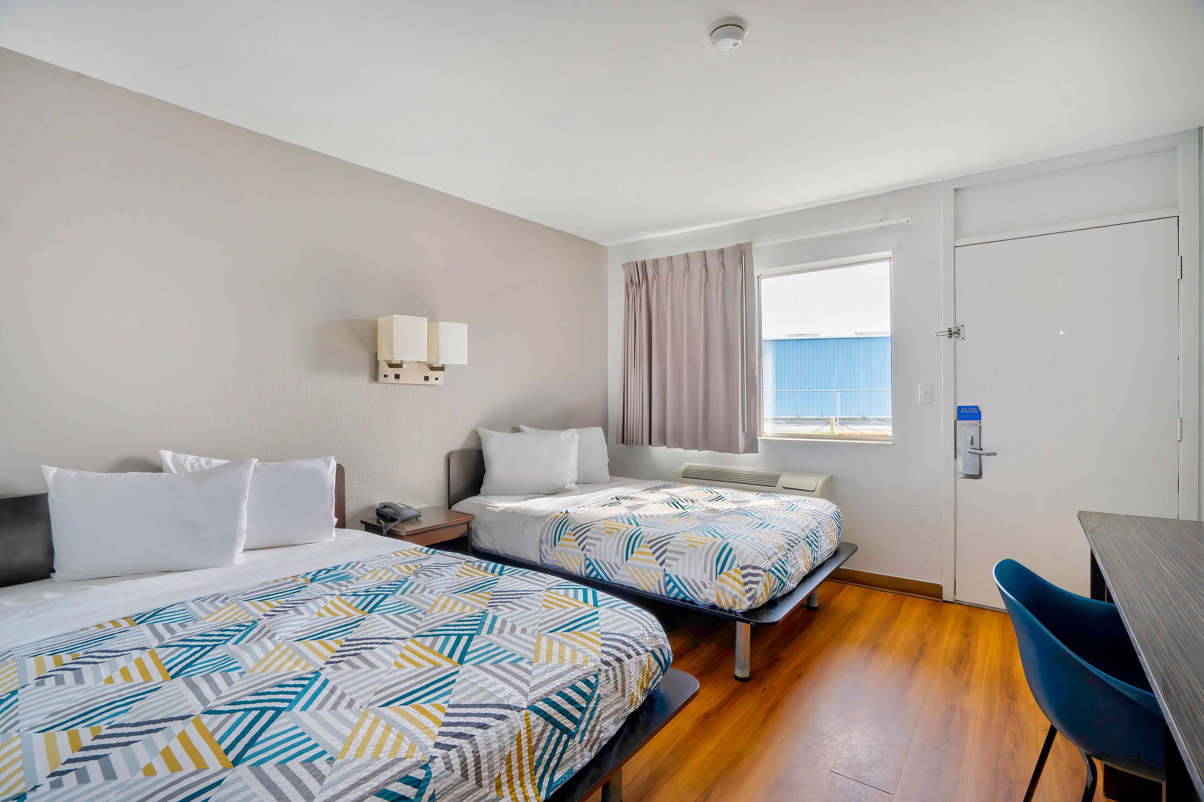 Quarto de hotel com duas camas, uma escrivaninha, uma cadeira, janela com cortinas, luminária na parede e porta com fechadura.
