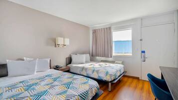 Quarto de hotel com duas camas, uma escrivaninha, uma cadeira, janela com cortinas, luminária na parede e porta com fechadura.