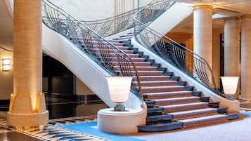 Uma escadaria imponente com grades de metal ricamente decoradas e carpete estampado, ladeada por grandes colunas e iluminada por arandelas.