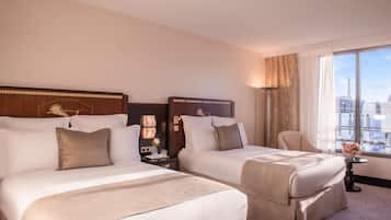 Quarto de hotel com duas camas, uma mesa de cabeceira com abajur, uma cadeira e vista para a cidade.