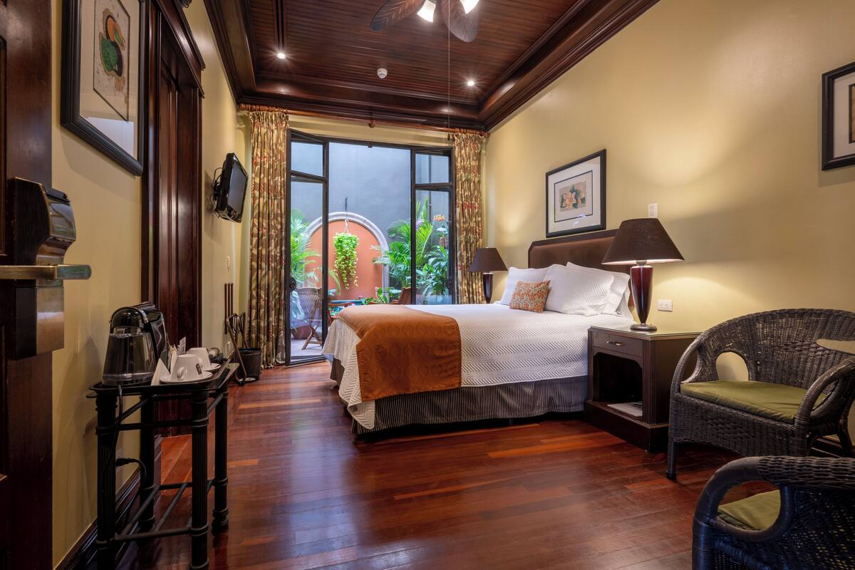 Quarto de hotel com uma cama grande, uma escrivaninha com abajur, uma cadeira de vime e vista para o verde lá fora.