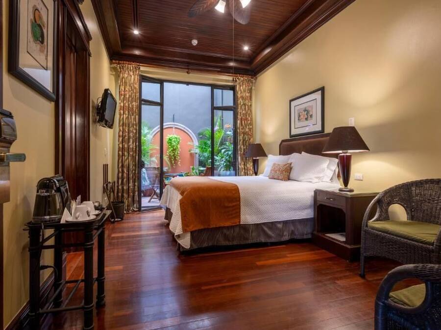 Quarto de hotel com uma cama grande, uma escrivaninha com abajur, cadeiras de vime e vista para o verde lá fora.
