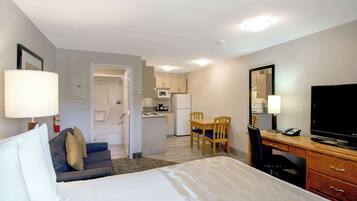 Quarto de hotel com cama, secretária, cadeira, televisão e cozinha compacta ao fundo.