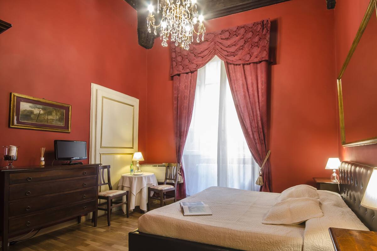 Quarto de hotel com cama, escrivaninha, cadeira, televisão, abajur e lustre.