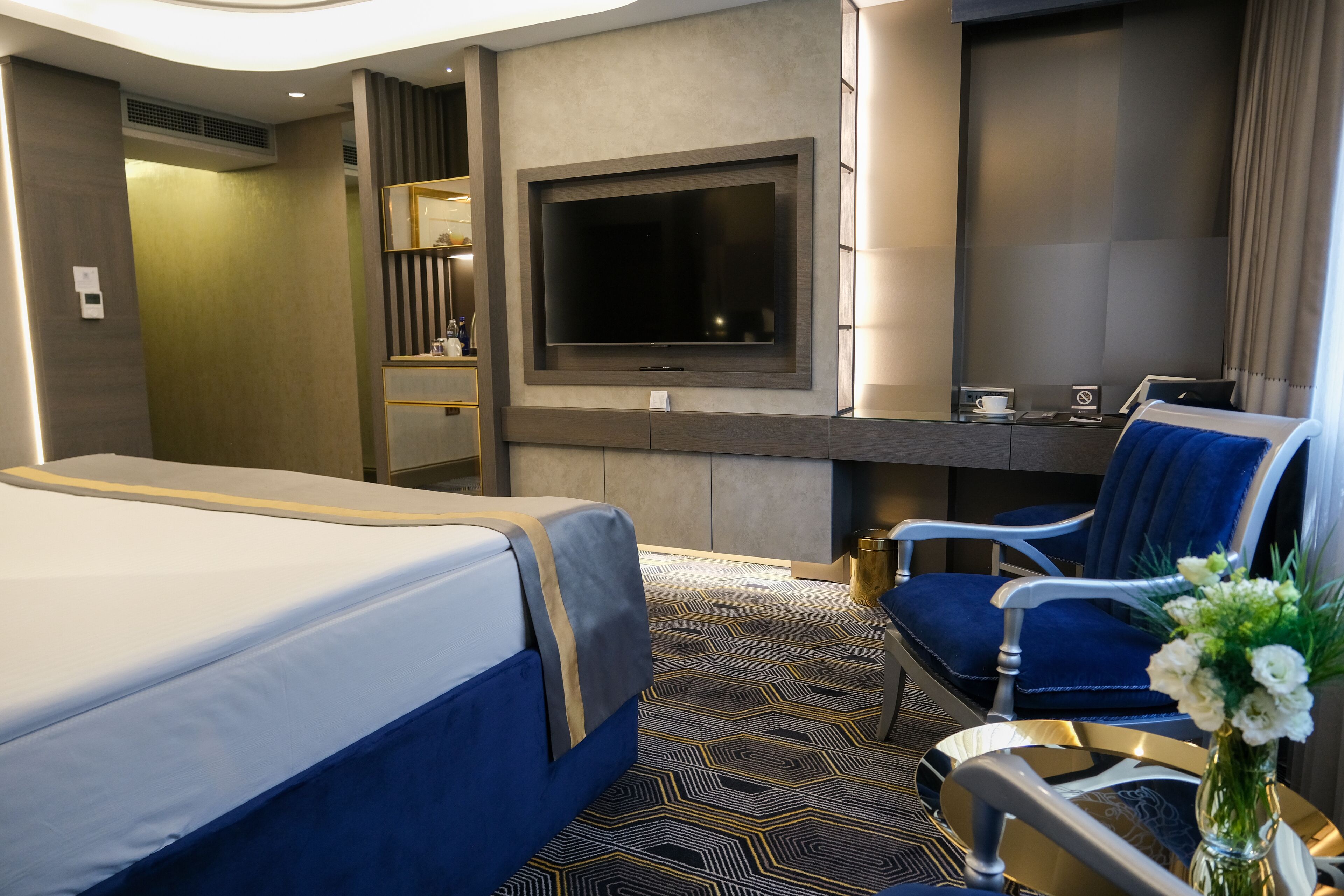 Quarto de hotel com uma cama grande, uma escrivaninha com televisão de tela plana, uma cadeira azul e um vaso com flores.