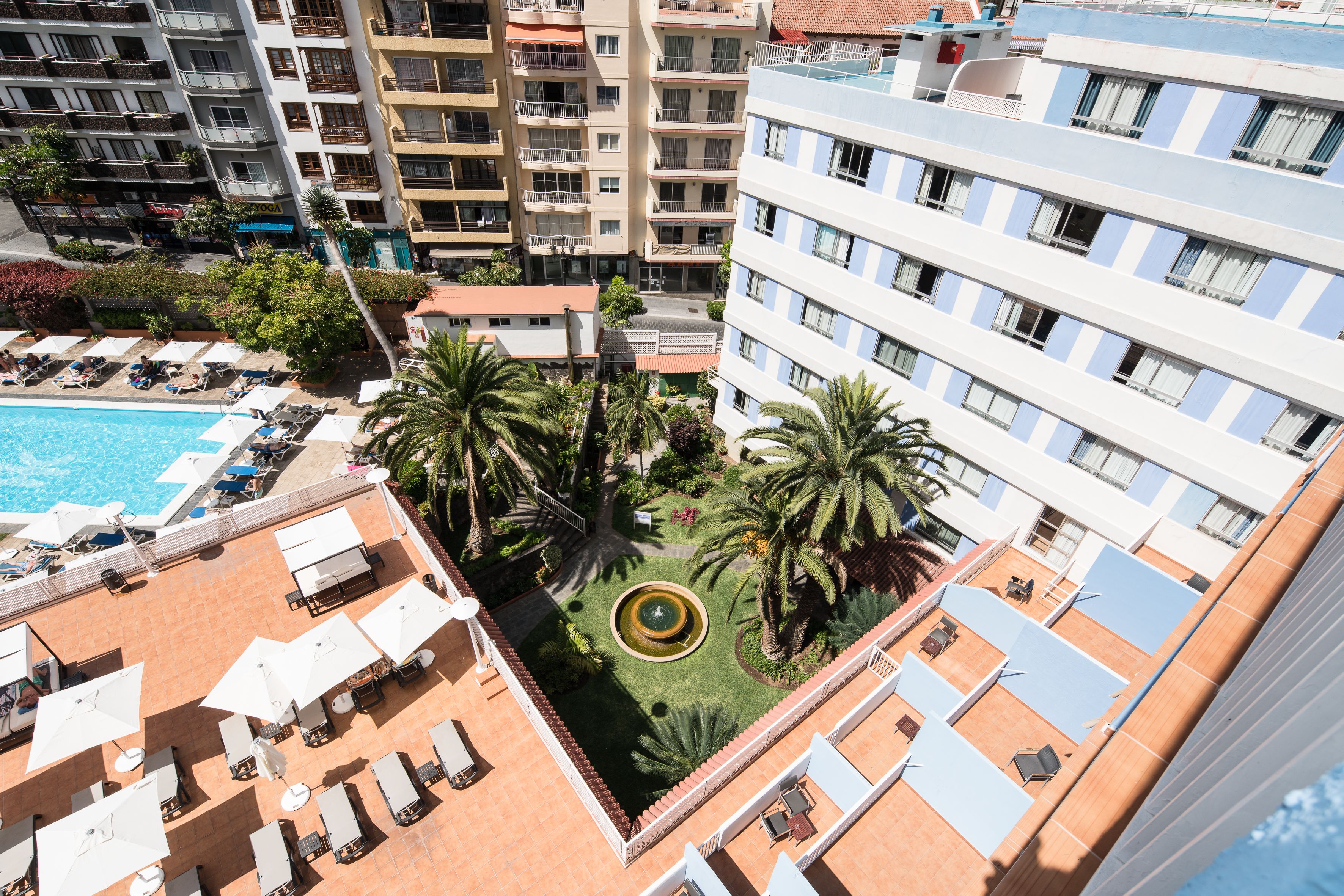 Área de piscina no terraço, com espreguiçadeiras e guarda-sóis, cercada por palmeiras e uma pequena fonte.